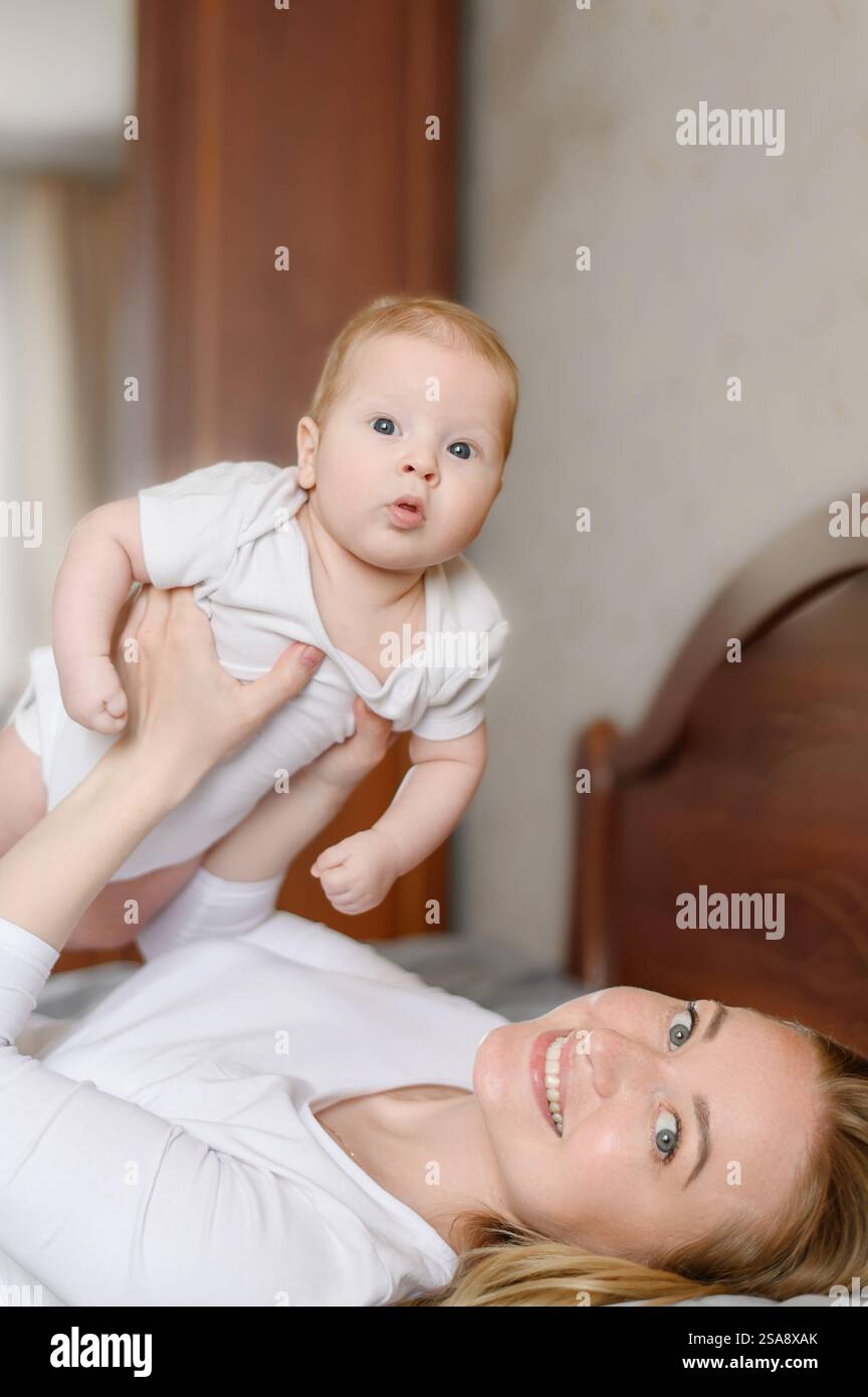 Happy woman lying on soft bed, tenderly holding her baby on her raised ...