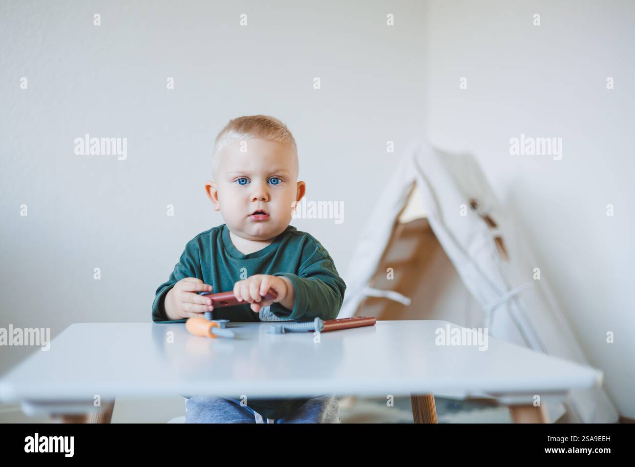 Toddler boy in a bright room sits at a children's table and plays with ...
