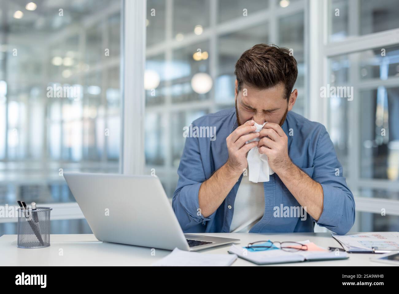 Sick man suffering at workplace, sneezing. Worker has runny nose and ...