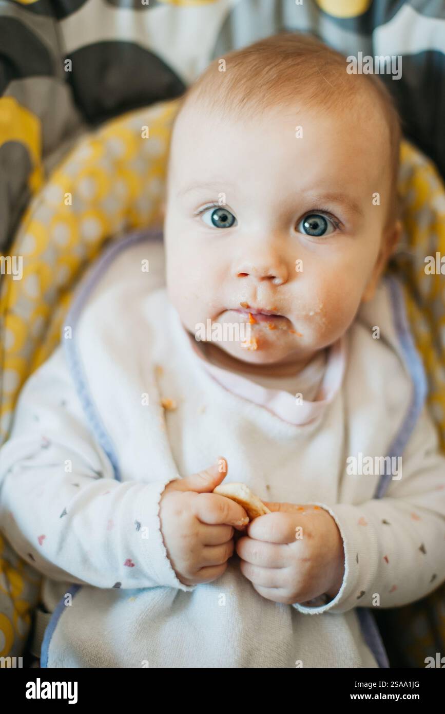 Cute Baby with Messy Face Eating Solid Food During Mealtime. Baby sitting in a high chair with ...