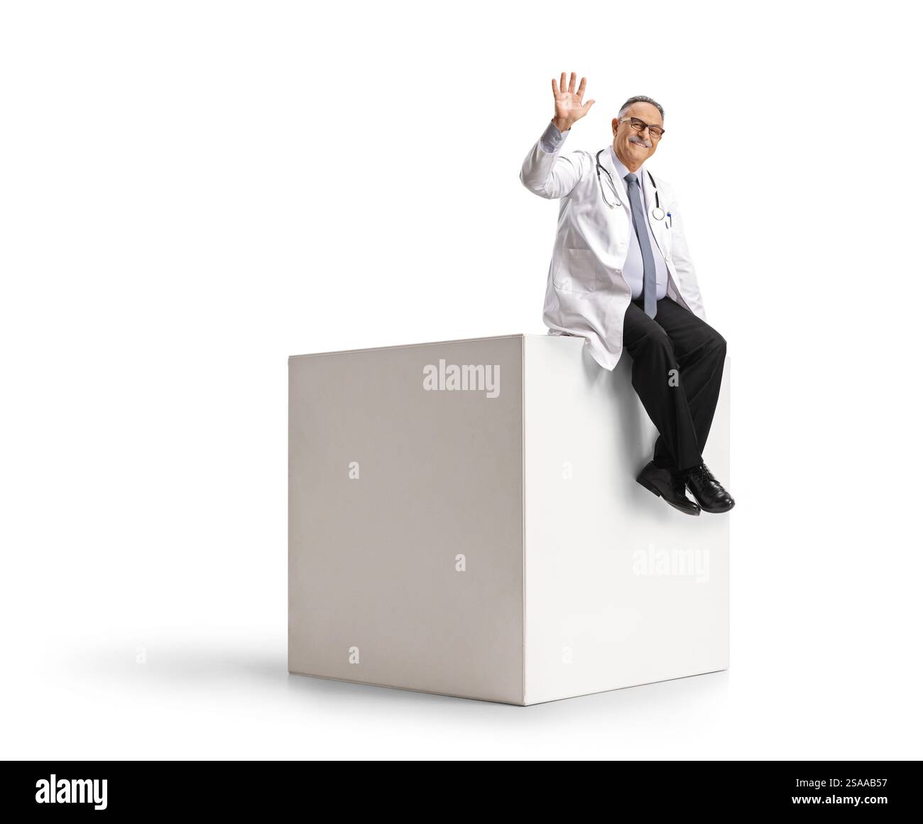 Mature male doctor sitting on a white cube and waving isolated on white ...