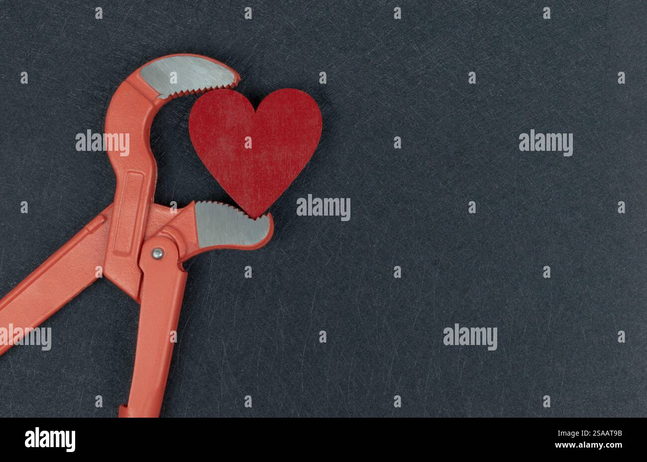 Red heart in jaws of a pipe wrench on black texture background. In jaws ...