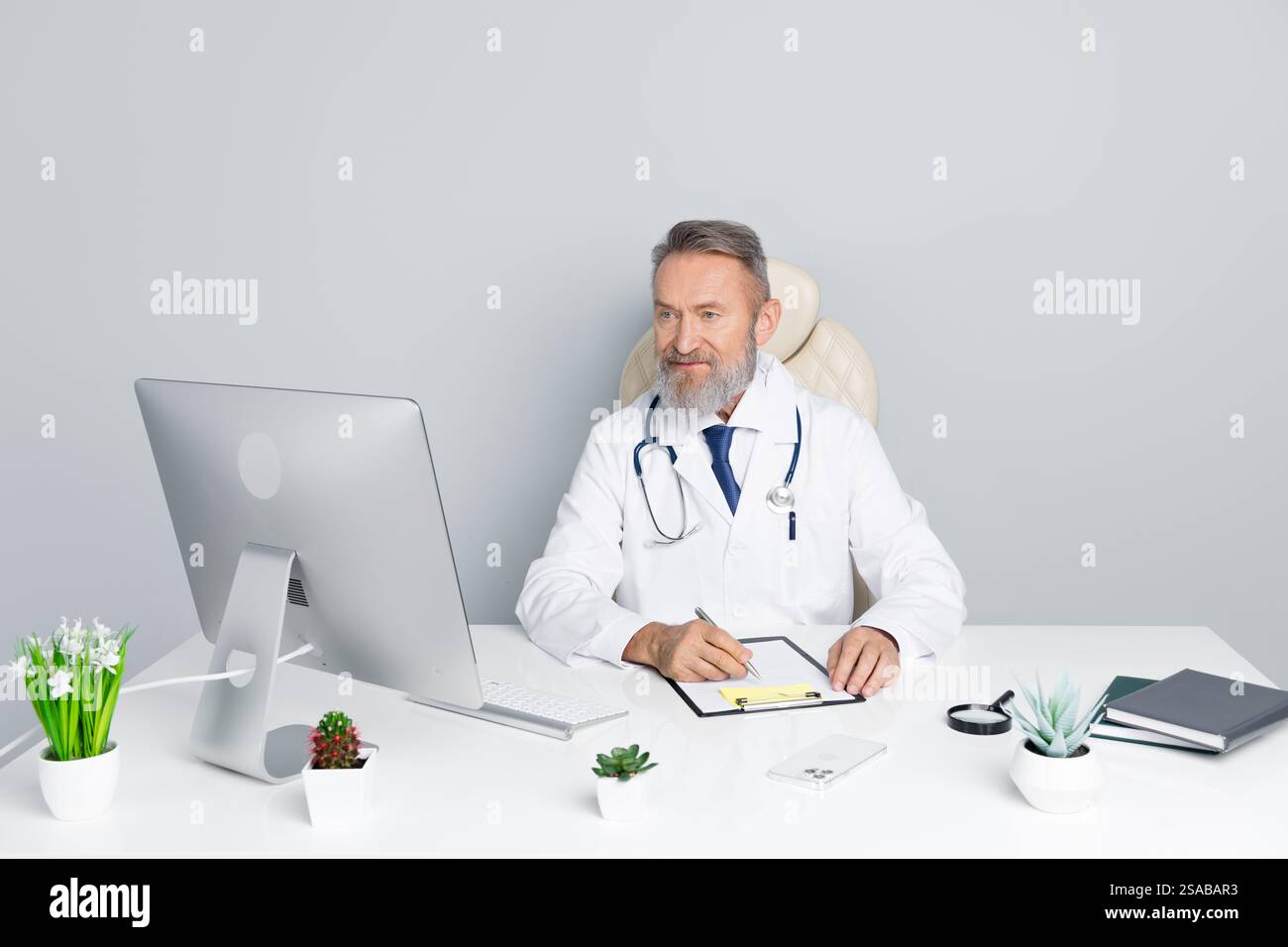 A senior male doctor writing notes at a modern desk with plants and ...