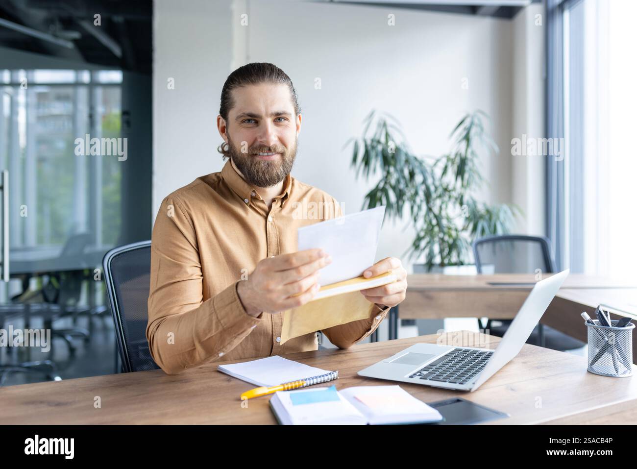 A smiling businessman sitting at a desk in an office, opening an envelope while using a laptop ...