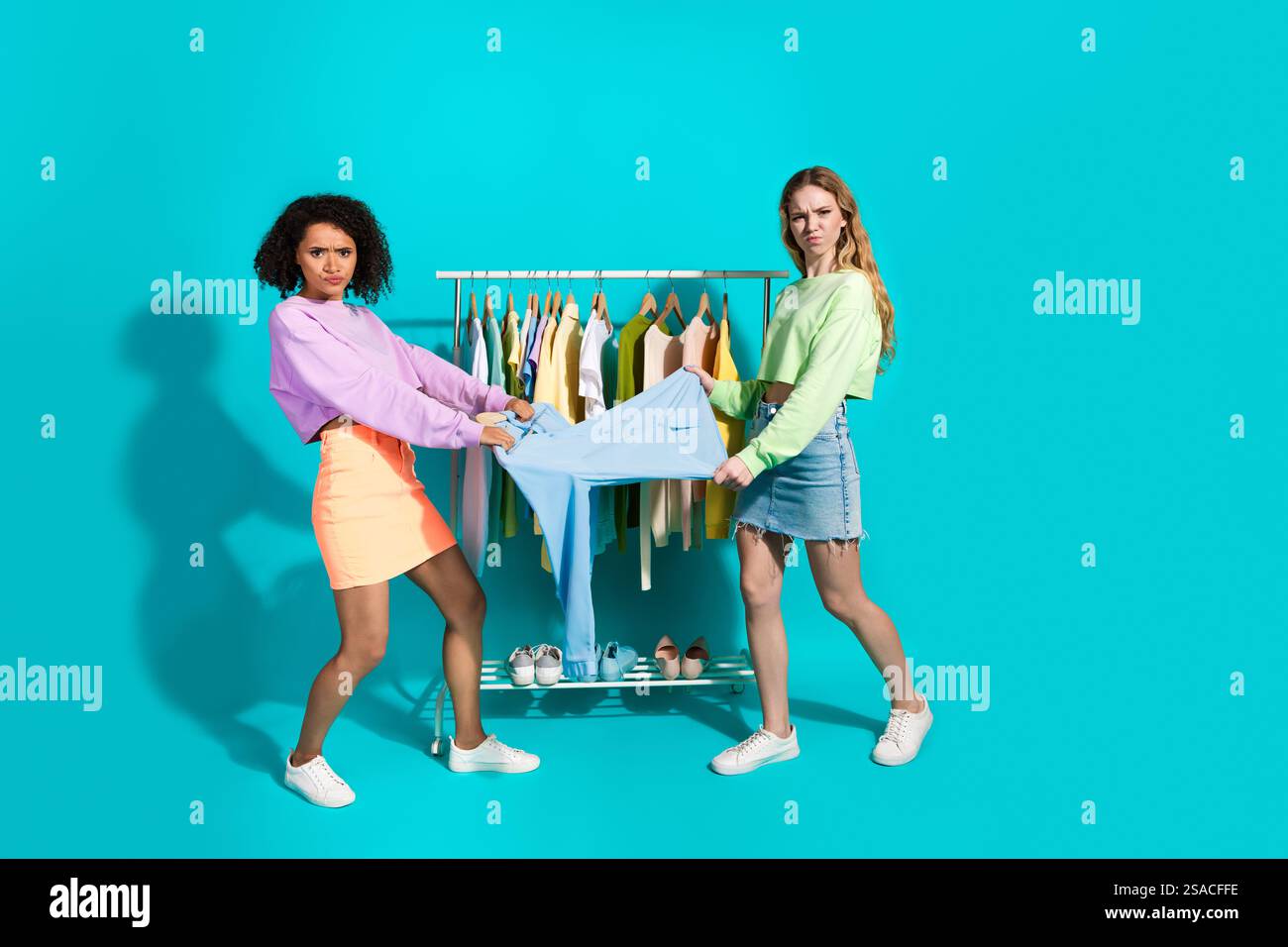 Two stylish young women pulling a garment with a wardrobe in a vibrant turquoise background ...