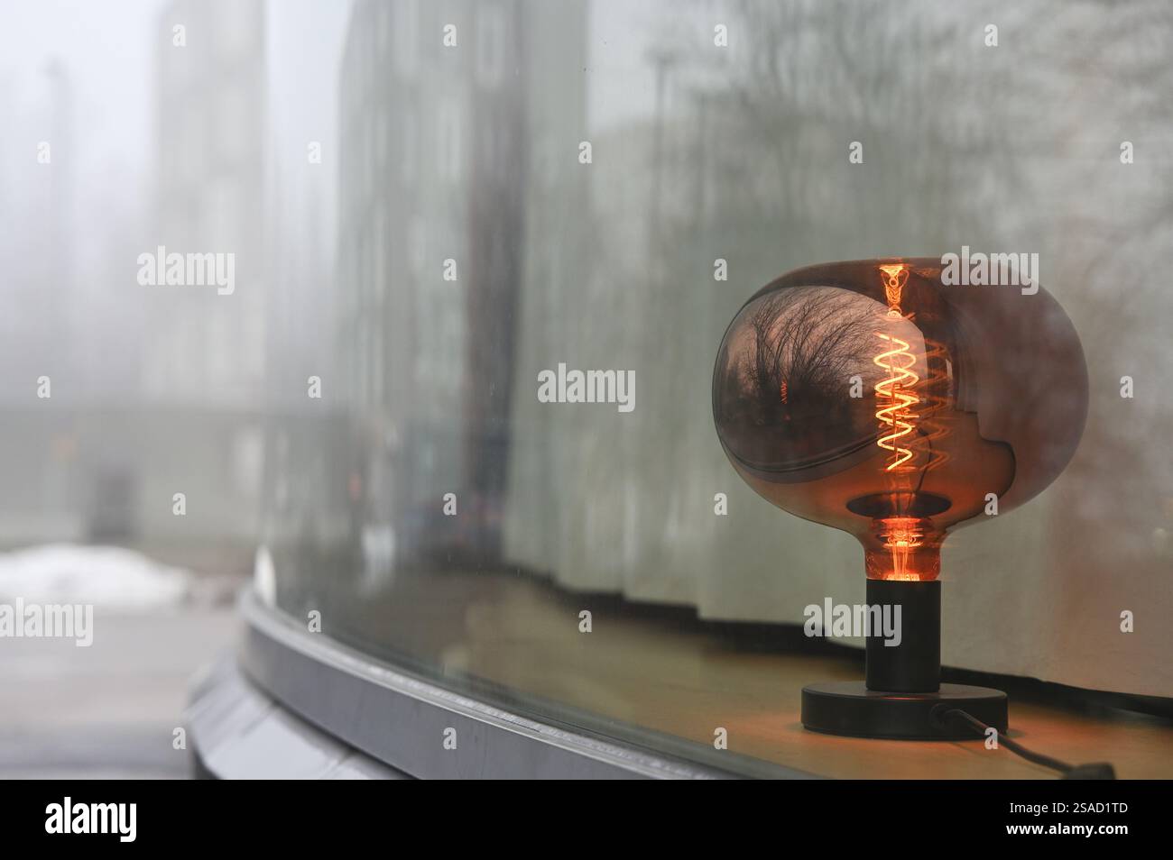 A warm glowing Edison-style lamp sits by a curved glass window, casting ...