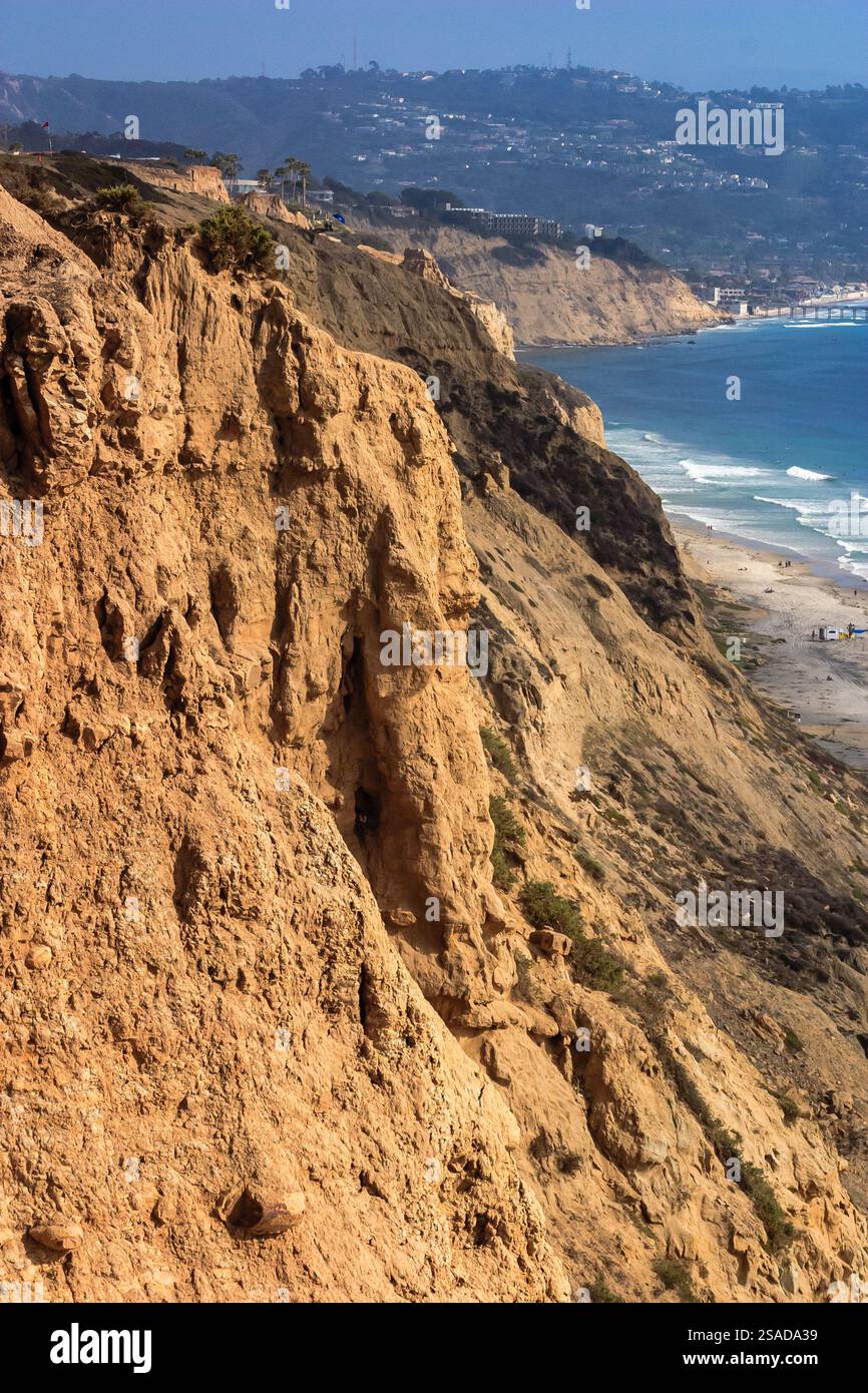 A rocky cliff overlooks the ocean. The beach is crowded with people ...