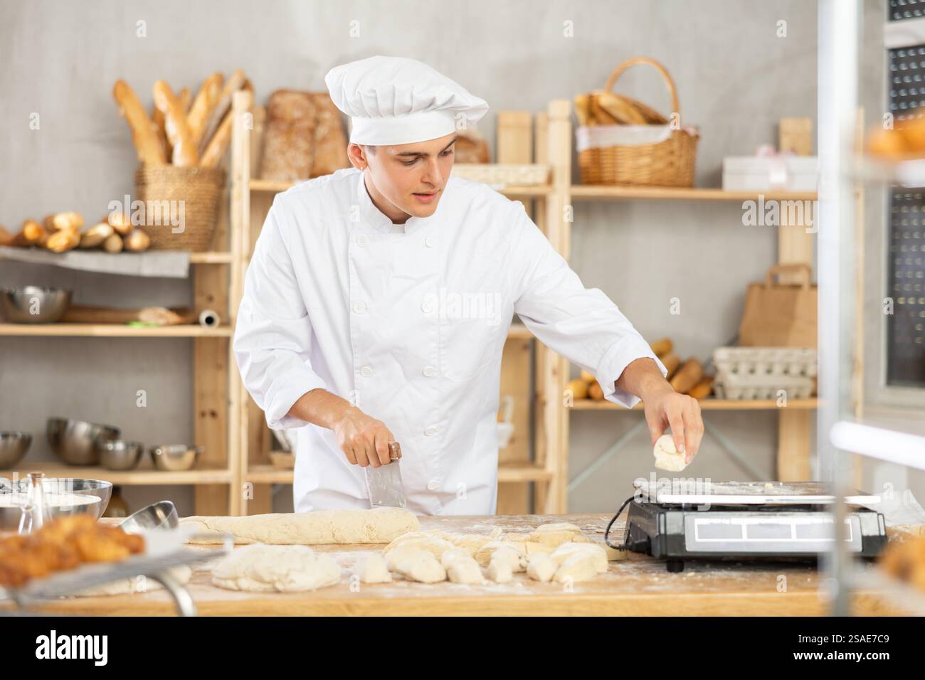 Young male baker cutting and weighing dough on scales in bakehouse ...