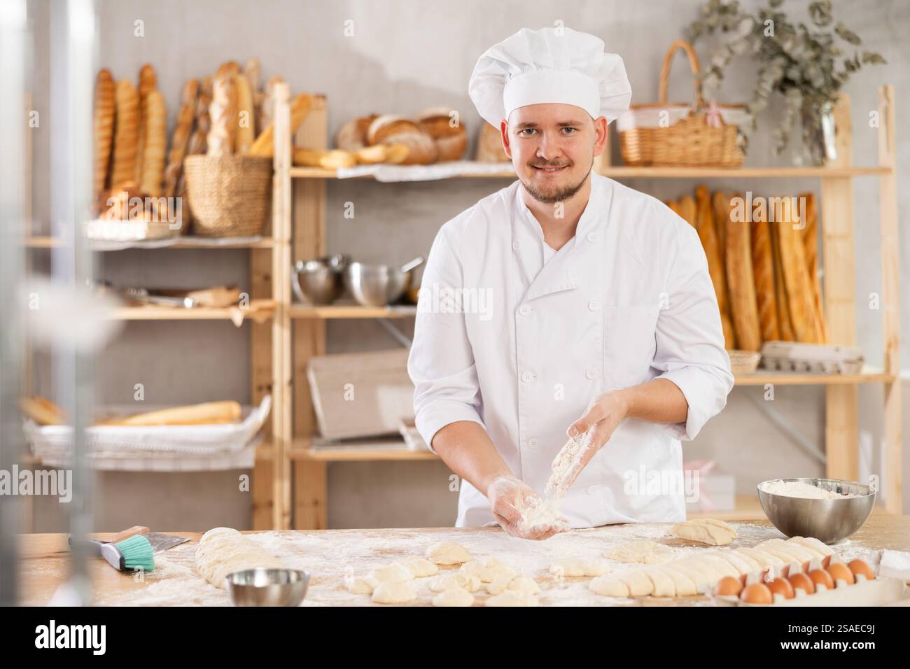 Professional baker stands at his work bench, kneading and shaping dough ...
