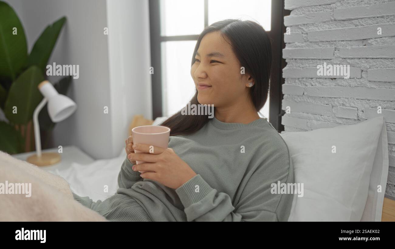 Woman relaxing indoors on bed holding mug looking content peaceful home interior setting asian ...