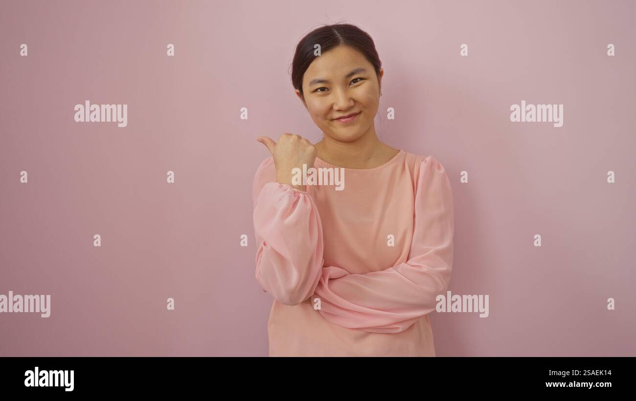 Woman smiling and pointing sideways with thumb against pink background ...