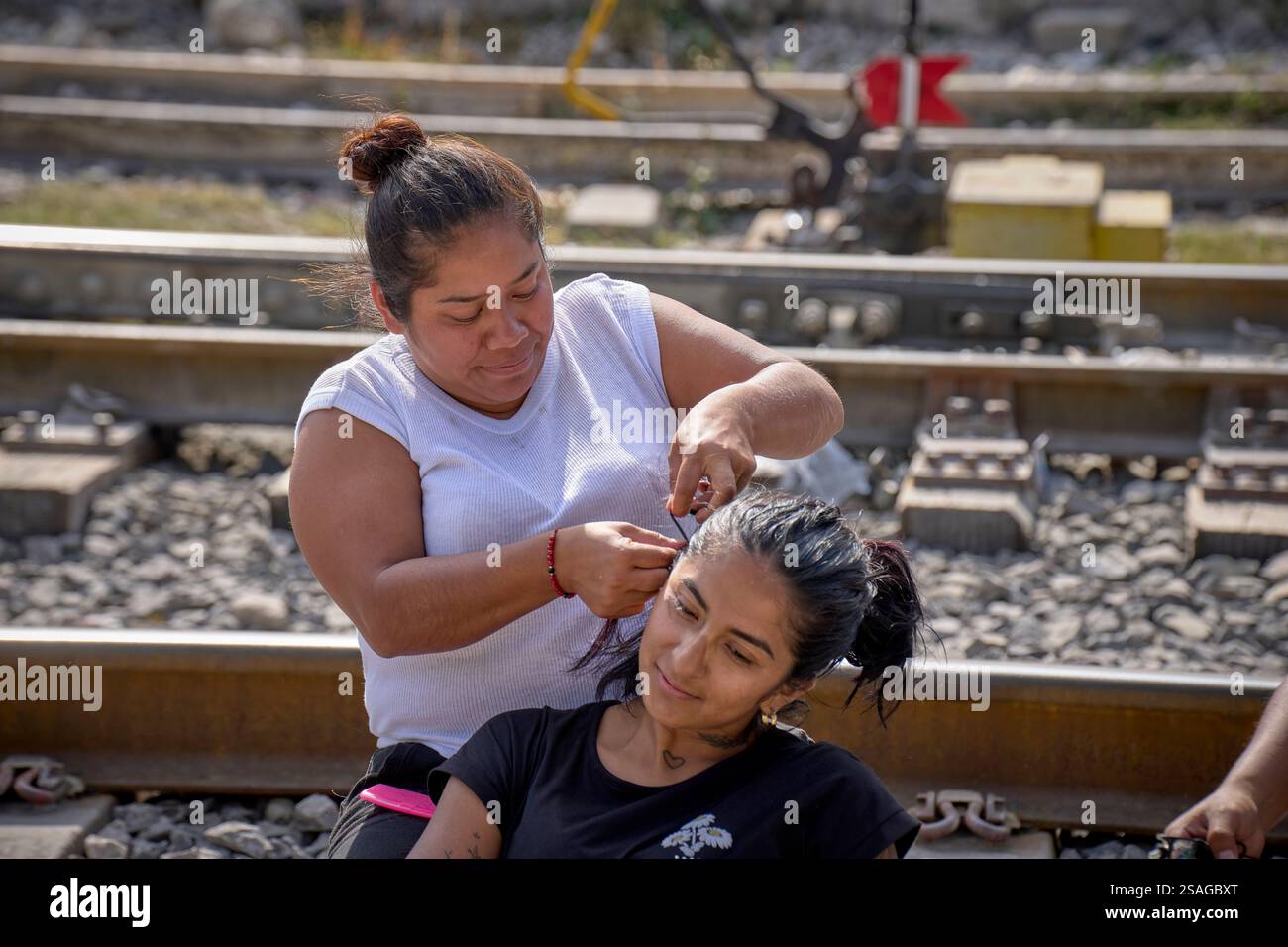 A migrant woman braids another woman's hair as they sit and wait along ...
