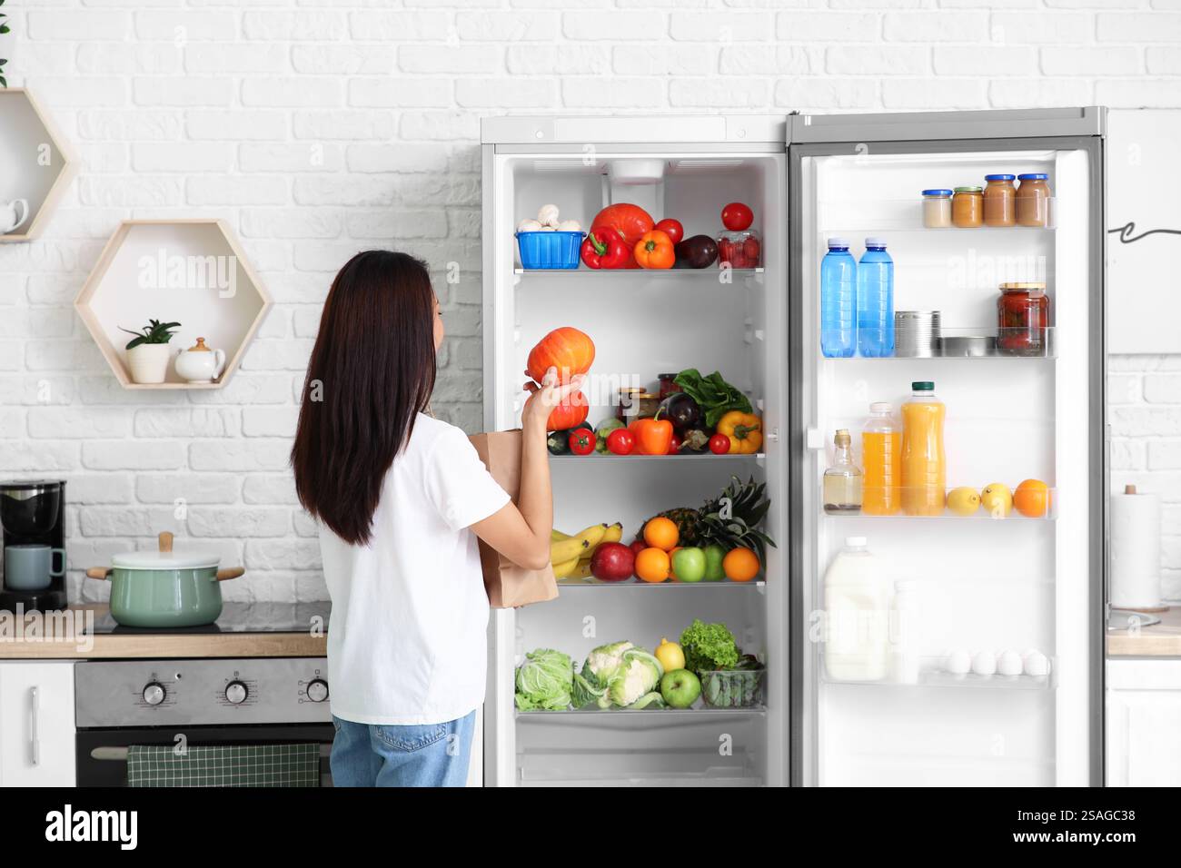 Young woman putting groceries into open fridge in kitchen, back view ...
