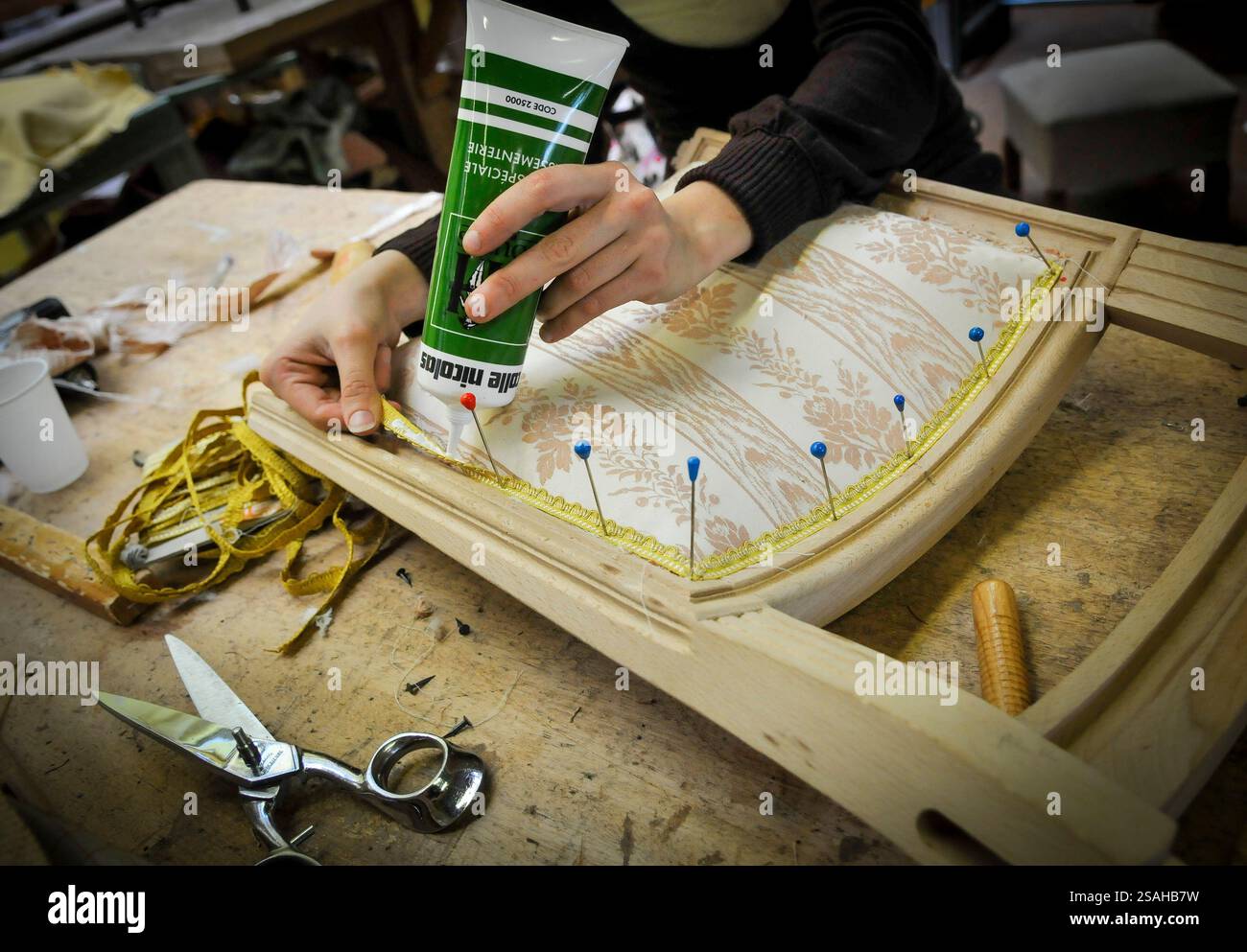 The upholsterer uses glue and pins to attach a braid to the back of an ...
