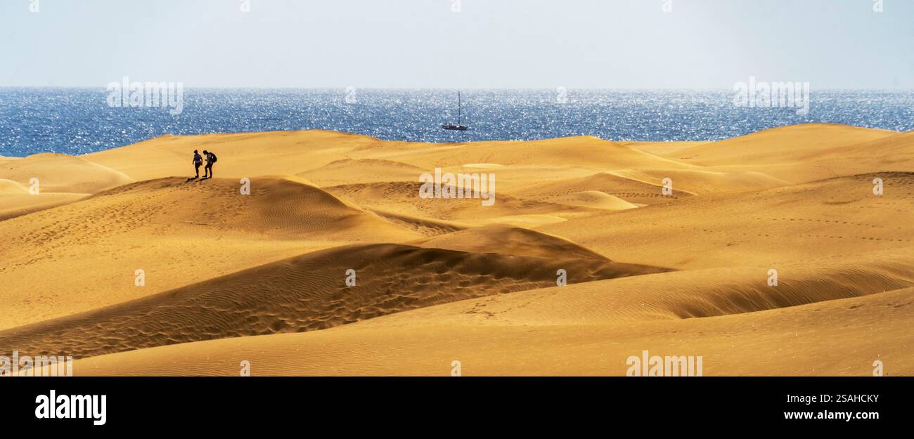 Two hikers walking on golden sand dunes with a shimmering ocean in the ...