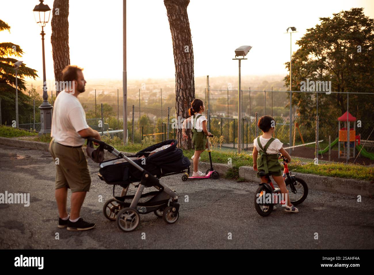 dad with baby in stroller and two bigger kids in bicycle and scooter walking towards playground ...
