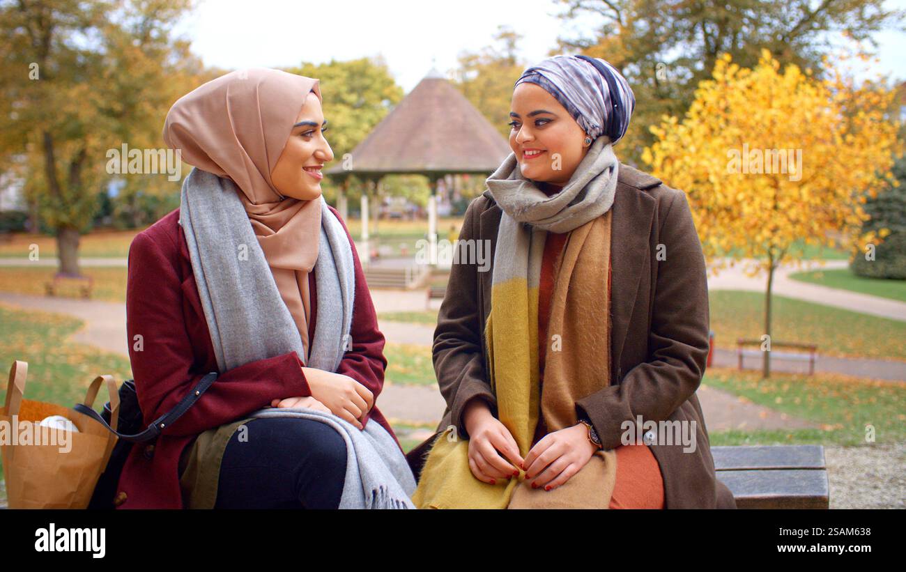 Two Young British Muslim Female Friends Sitting On Bench In Autumn Park ...
