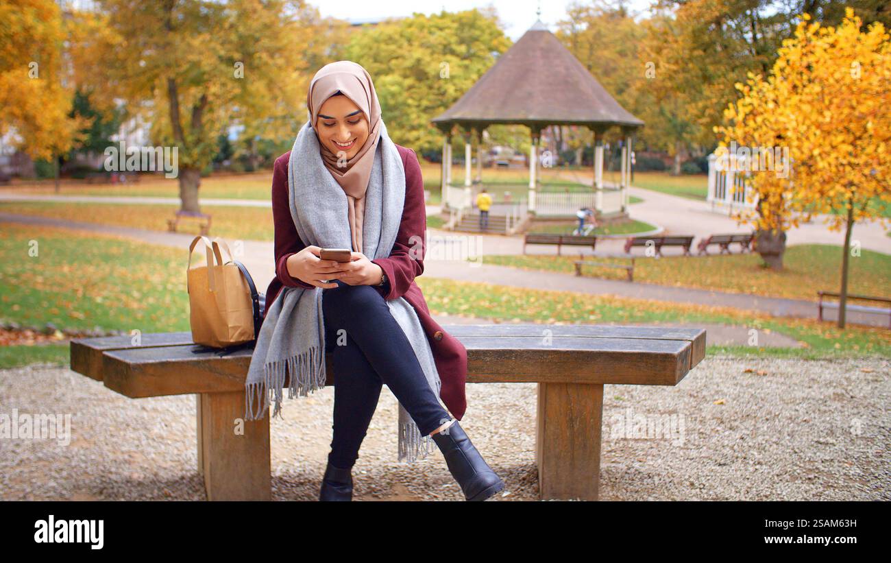 British Muslim Woman Sitting On Bench Using Mobile Phone To Check ...