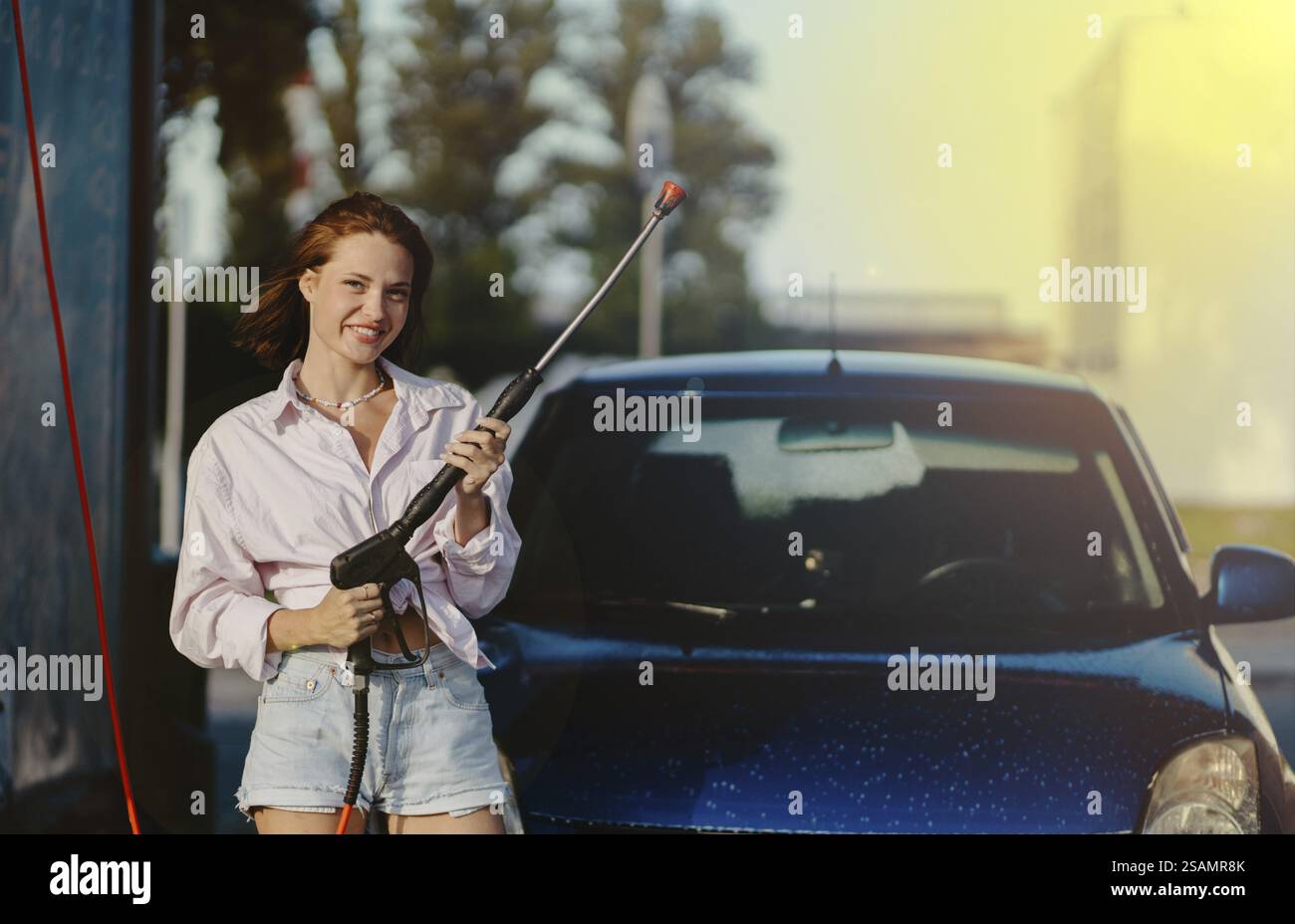 Woman holding a pressure washer stands near a blue car, smiling in the ...