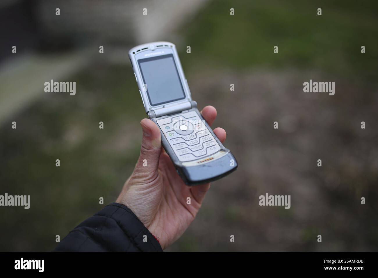 A hand holds an open gray flip phone outdoors against a grassy backdrop ...