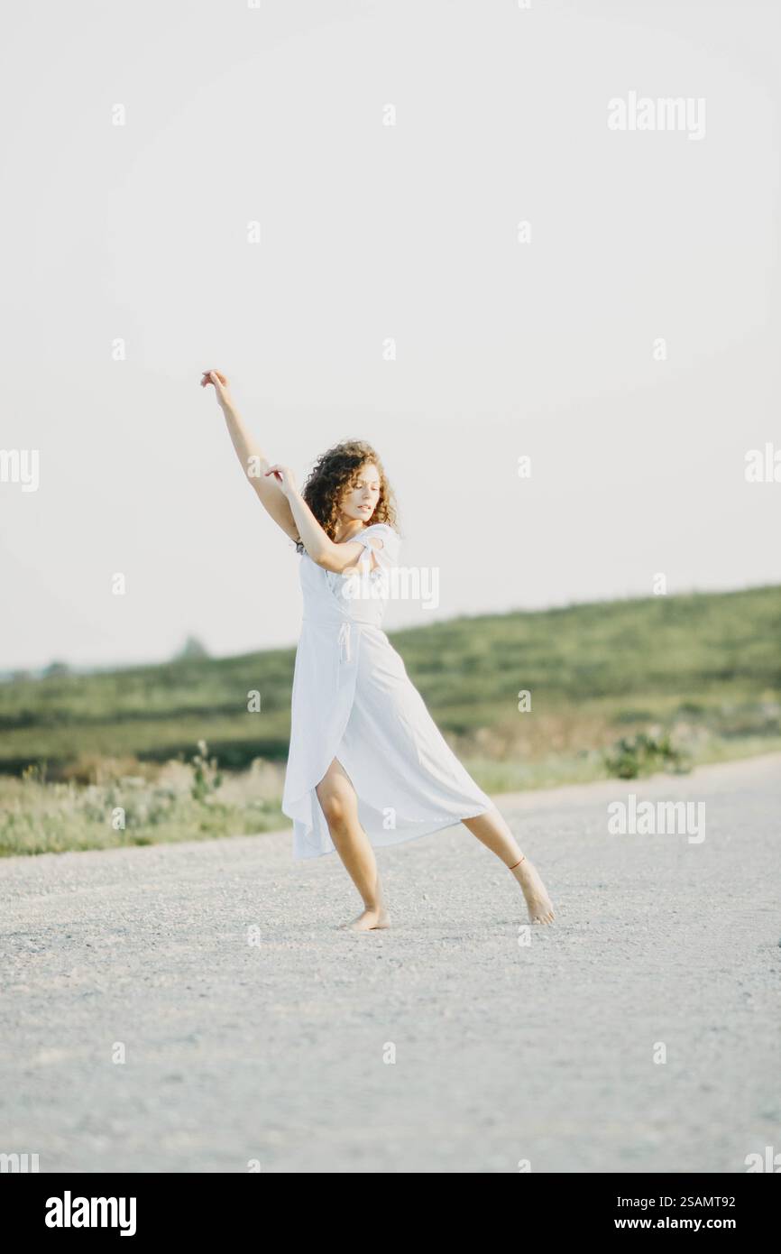 Woman dancing gracefully in a white dress on a dusty path near a green ...
