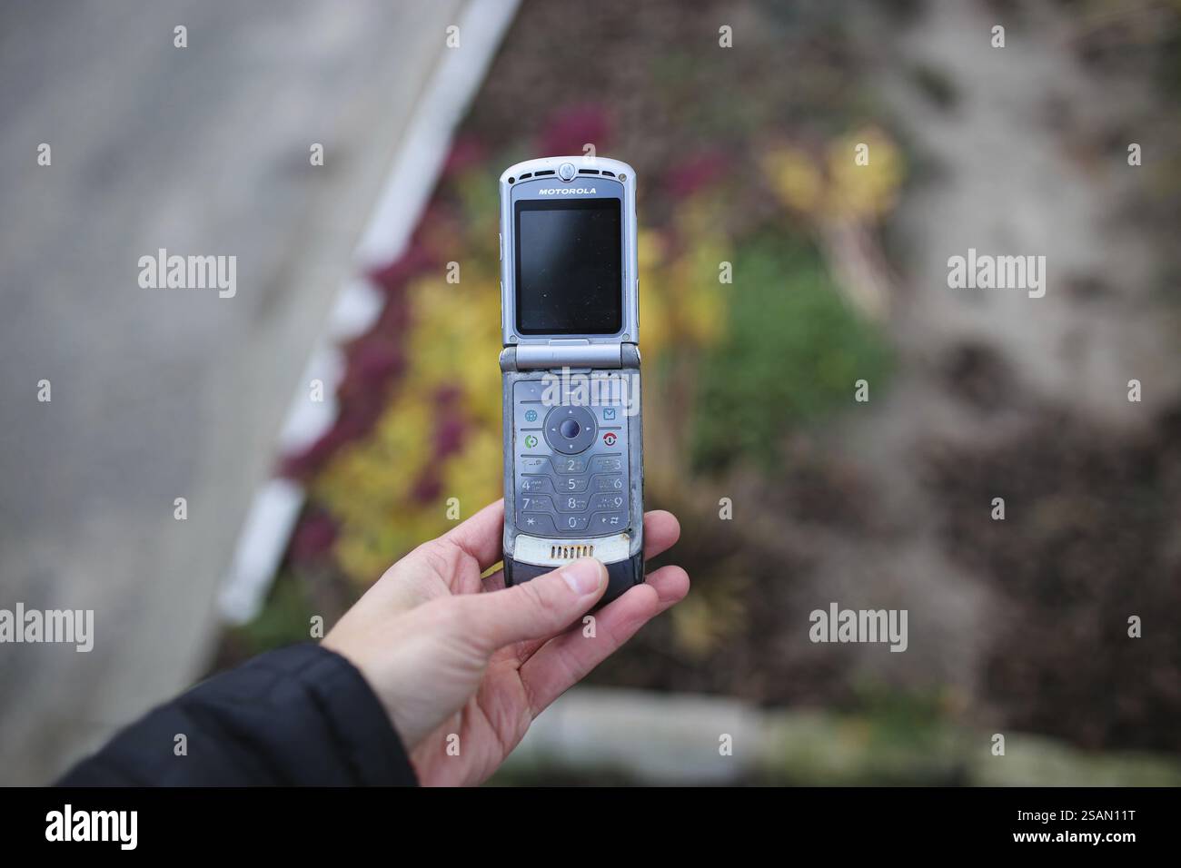 An open gray flip phone held in a hand outdoors with colorful ...