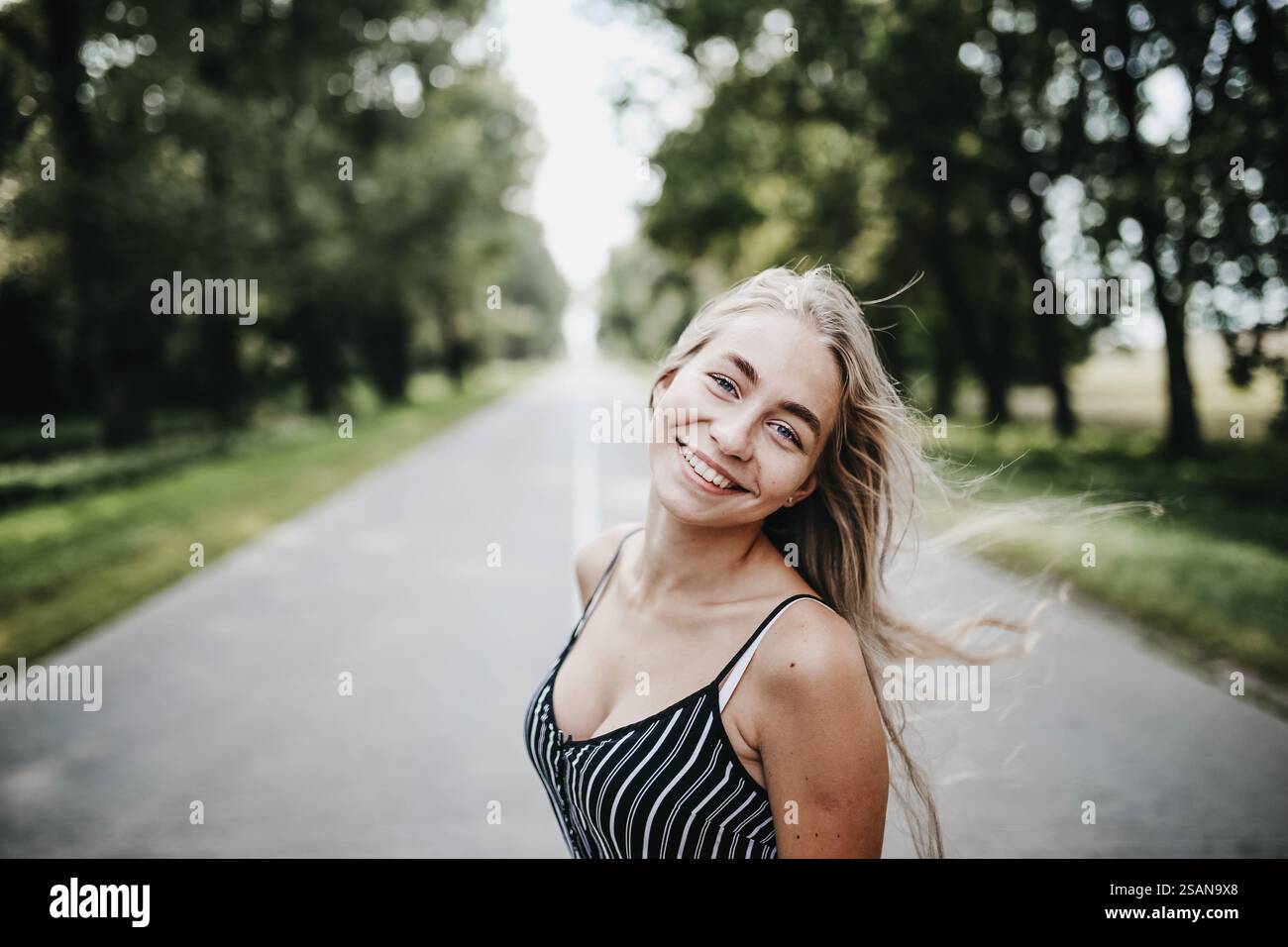 Woman with a wide smile on a tree-lined road, her hair carried by the ...