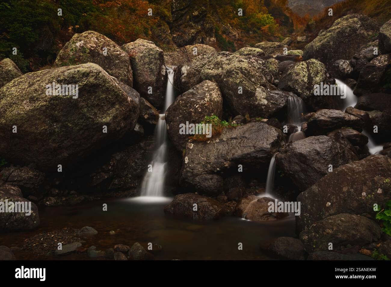 Cascading waterfalls flow over moss-covered boulders surrounded by autumn colors, creating a ...