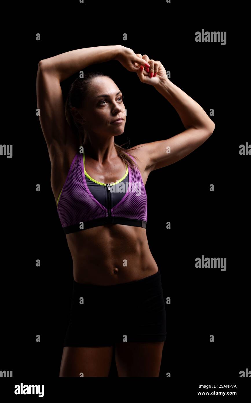 Confident fit woman stretching her arms while wearing sporty attire on a black background ...