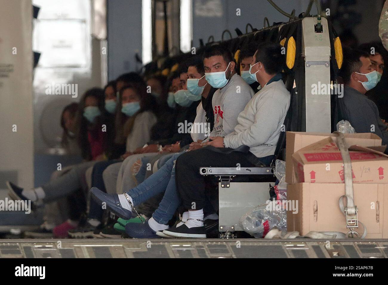 Migrants wearing face masks and shackles on their hands and feet sit on ...
