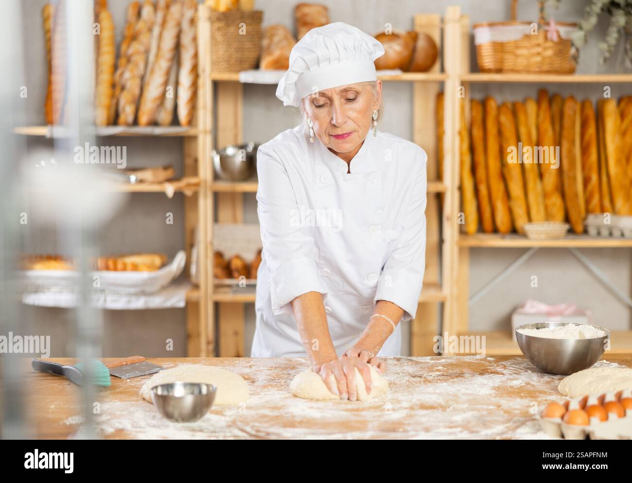 Senior woman works in bakery as baker, kneads dough, works with flour ...