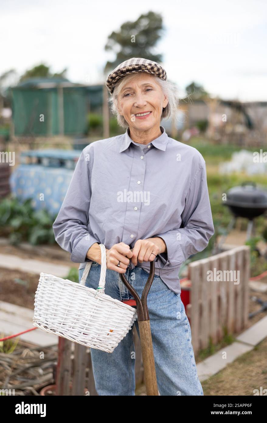 Happy mature female holding wicker basket and handle of shovel while ...