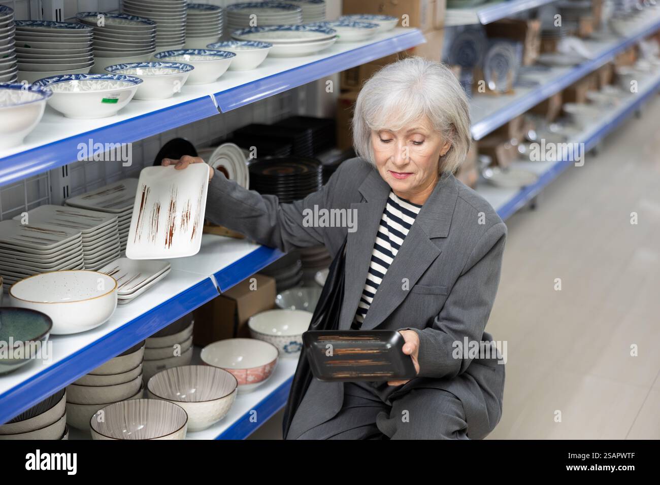 Enthusiastic senior lady choosing square ceramic plates at Asian store ...