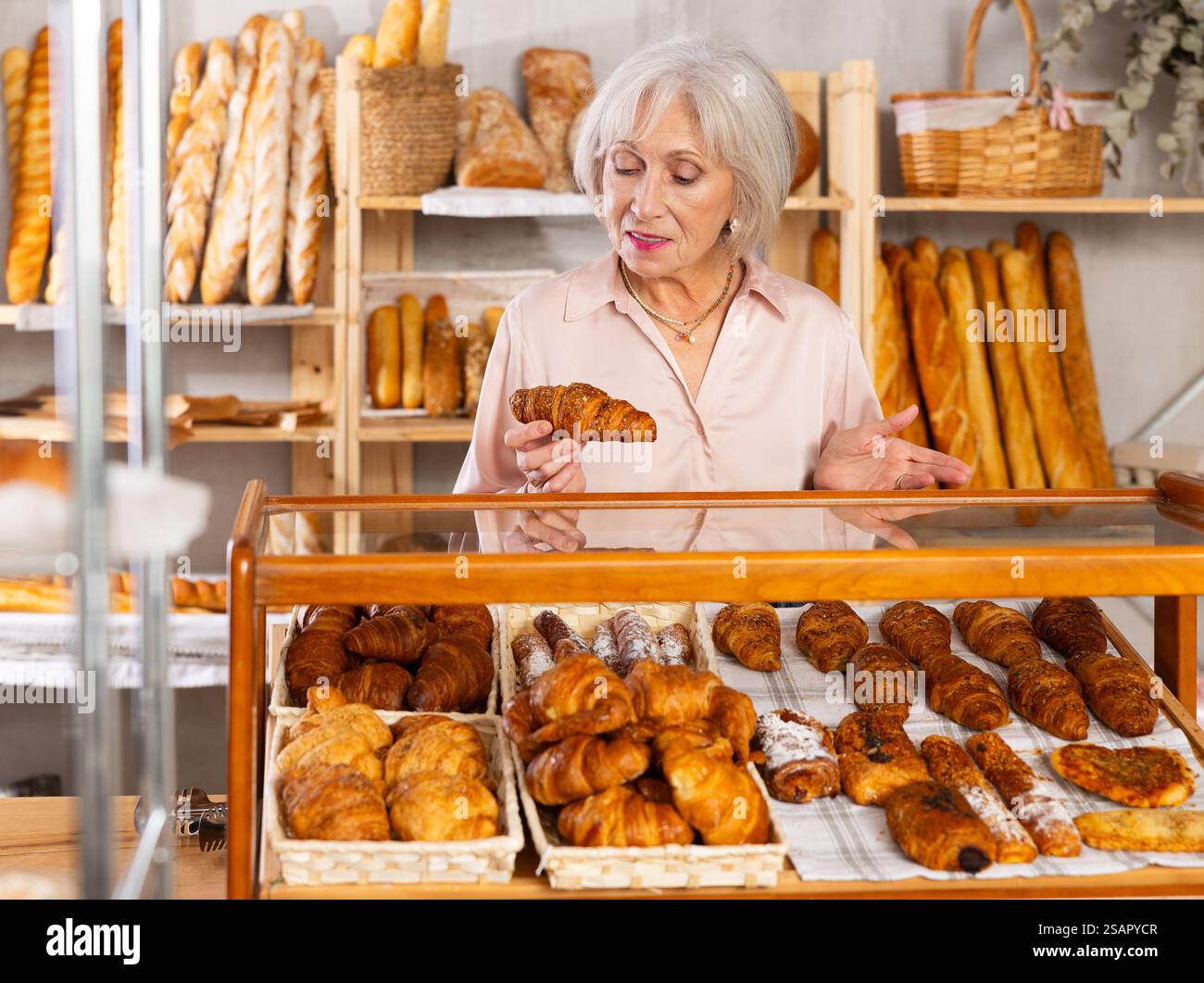 Senior female customer stands with croissant in hands near window of ...