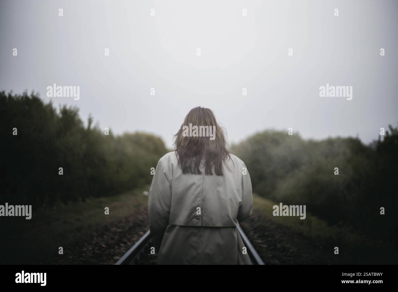 A woman stands alone on train tracks under an overcast sky, creating a ...