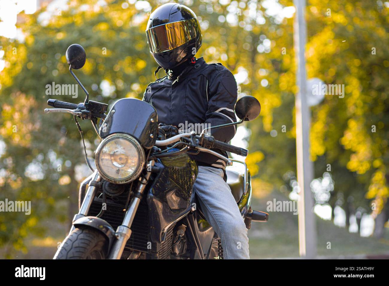 A motorcyclist wearing a helmet stands with a bike on a leafy street ...