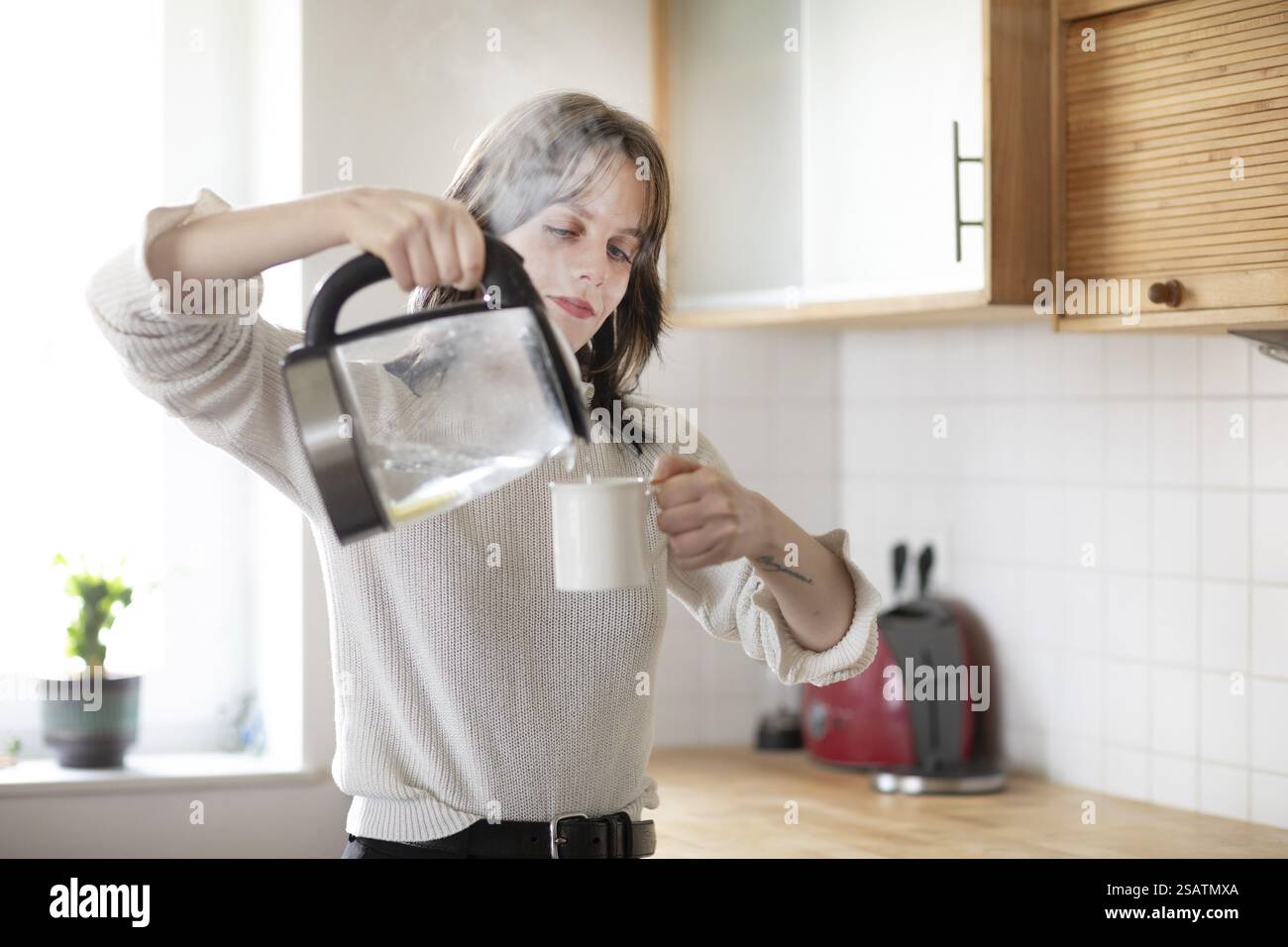 Woman pouring water into a cup in a bright kitchen with kettle, energy ...