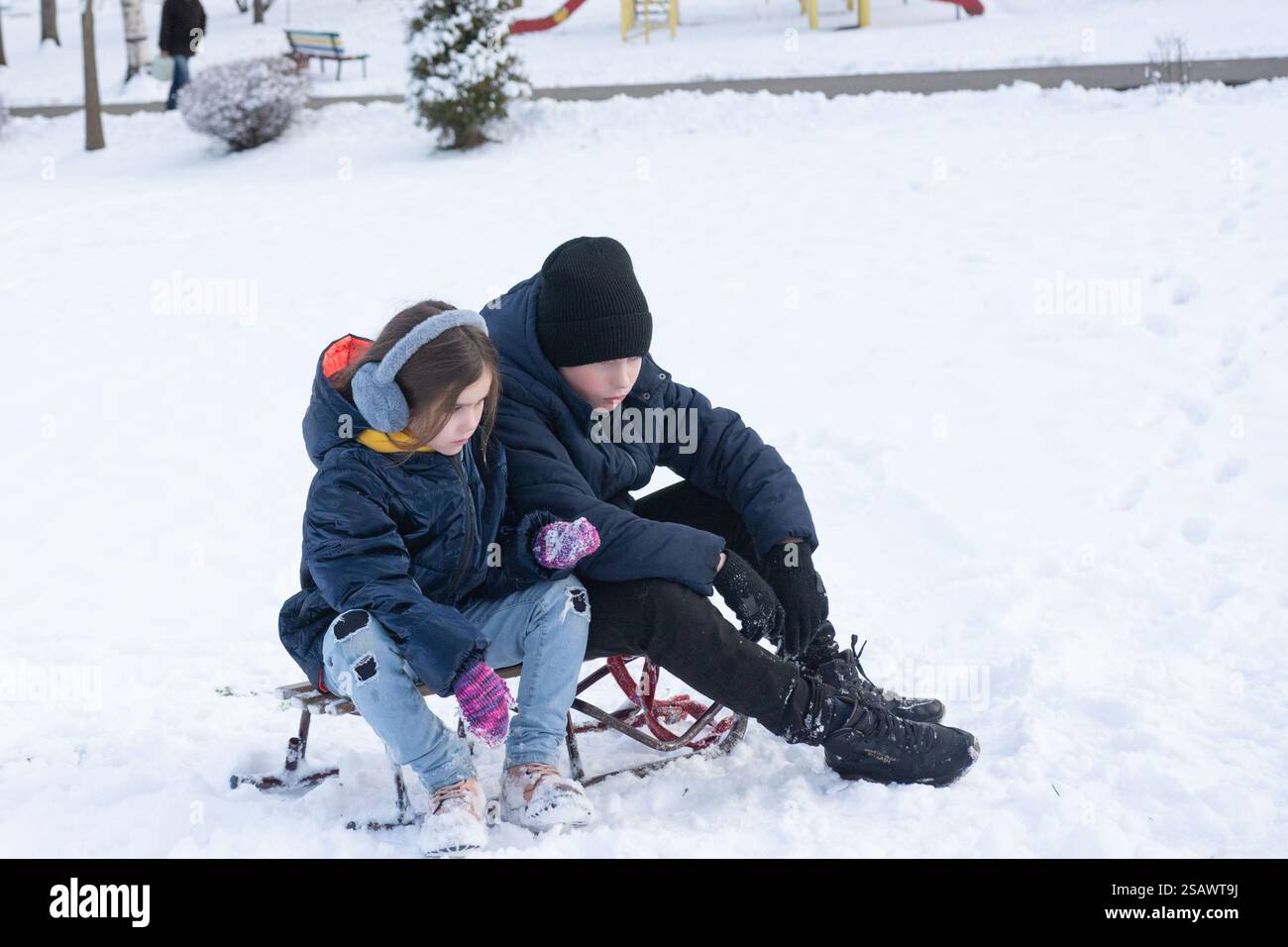 Two children, a boy and a girl, sit on a sled in the snow. They are ...