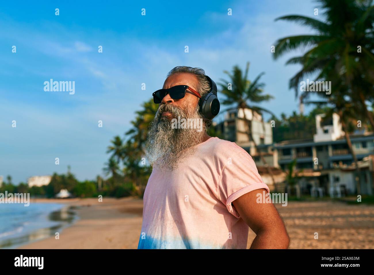 Grey-bearded senior man enjoying music on tropical beach. Stylish elder ...