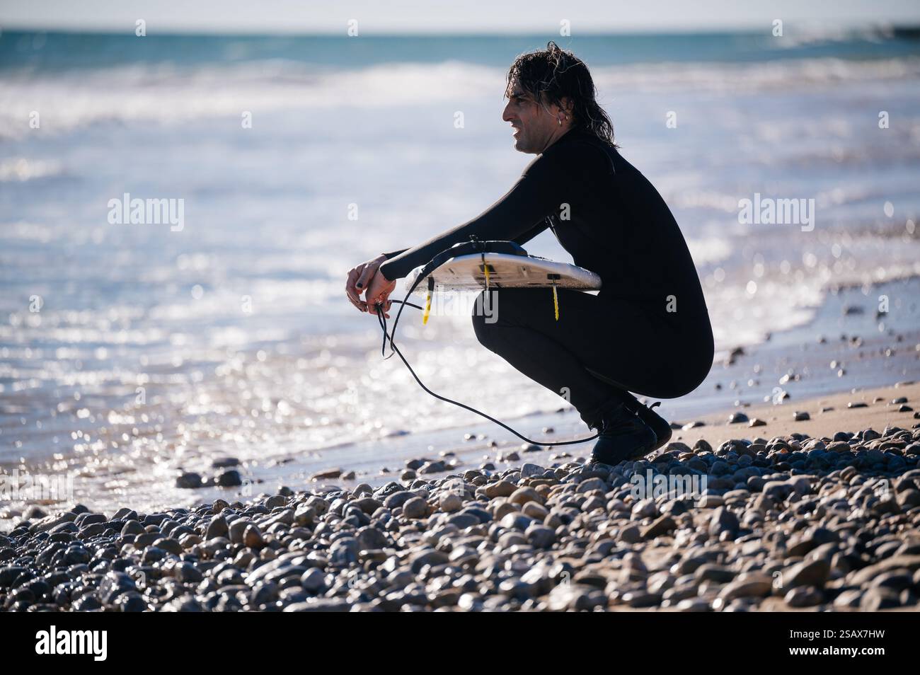 Surfer crouching on the beach holding his surfboard Stock Photo - Alamy