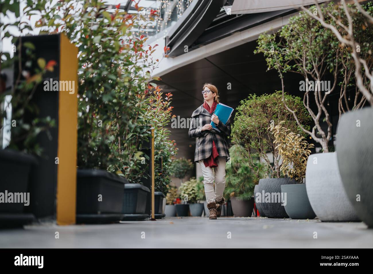 Professional woman walking by outdoor potted plants carrying folders ...