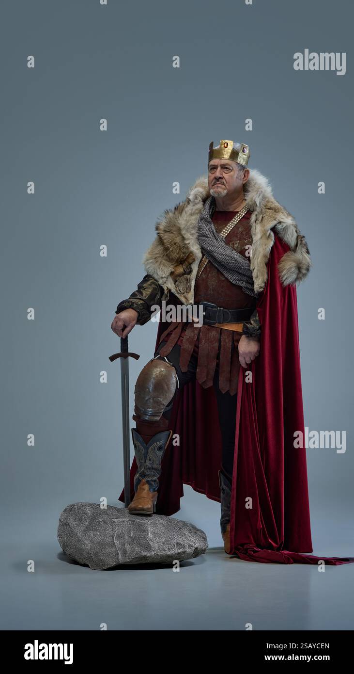 Senior serious man as medieval king standing on stone, leaning on sword ...