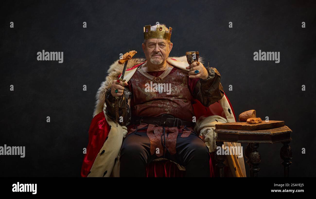 Senior man, medieval king in period attire sitting on throne with meat ...