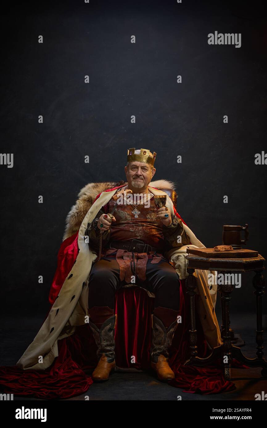 Senior man, medieval king in period attire sitting on throne with meat ...