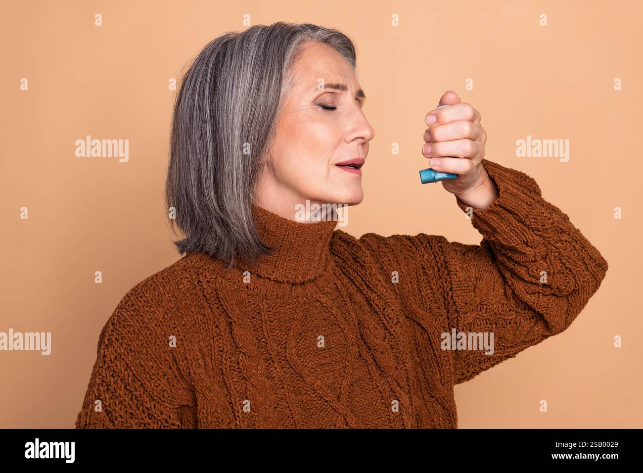 Elderly woman with grey hair using inhaler against beige background in ...