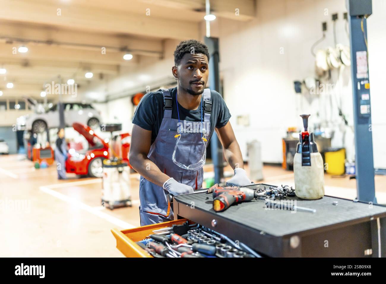 Young african car mechanic pushing a tool trolley in a busy workshop ...