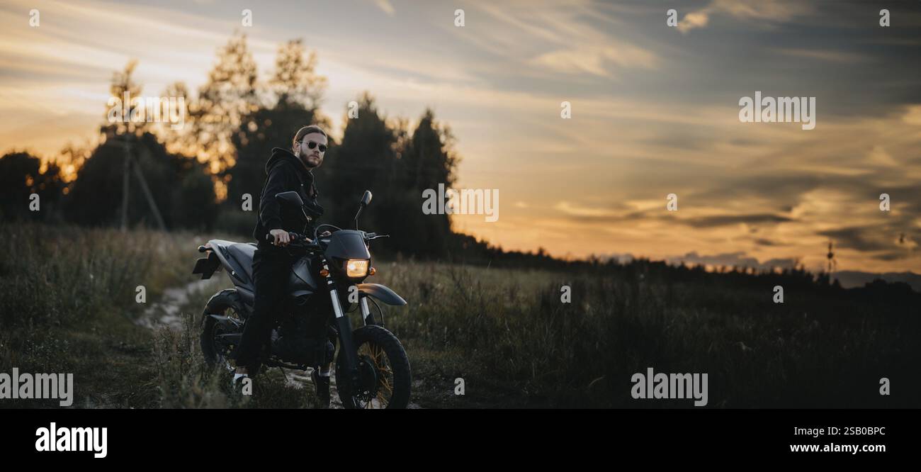 Man beside a motorcycle in an open field at sunset, sky displaying ...