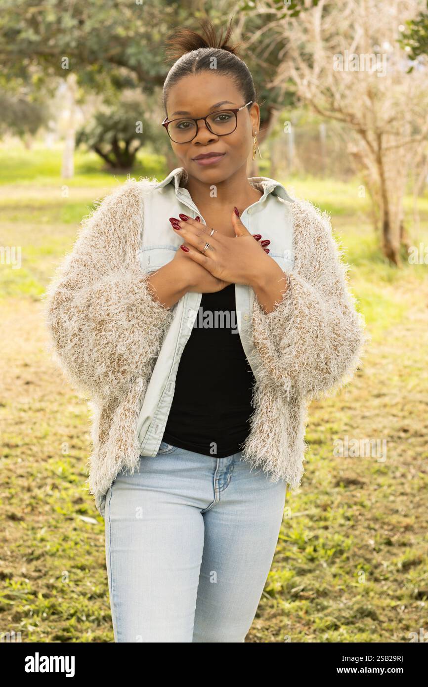 A black woman stands outdoors with her hands gently placed on her chest ...