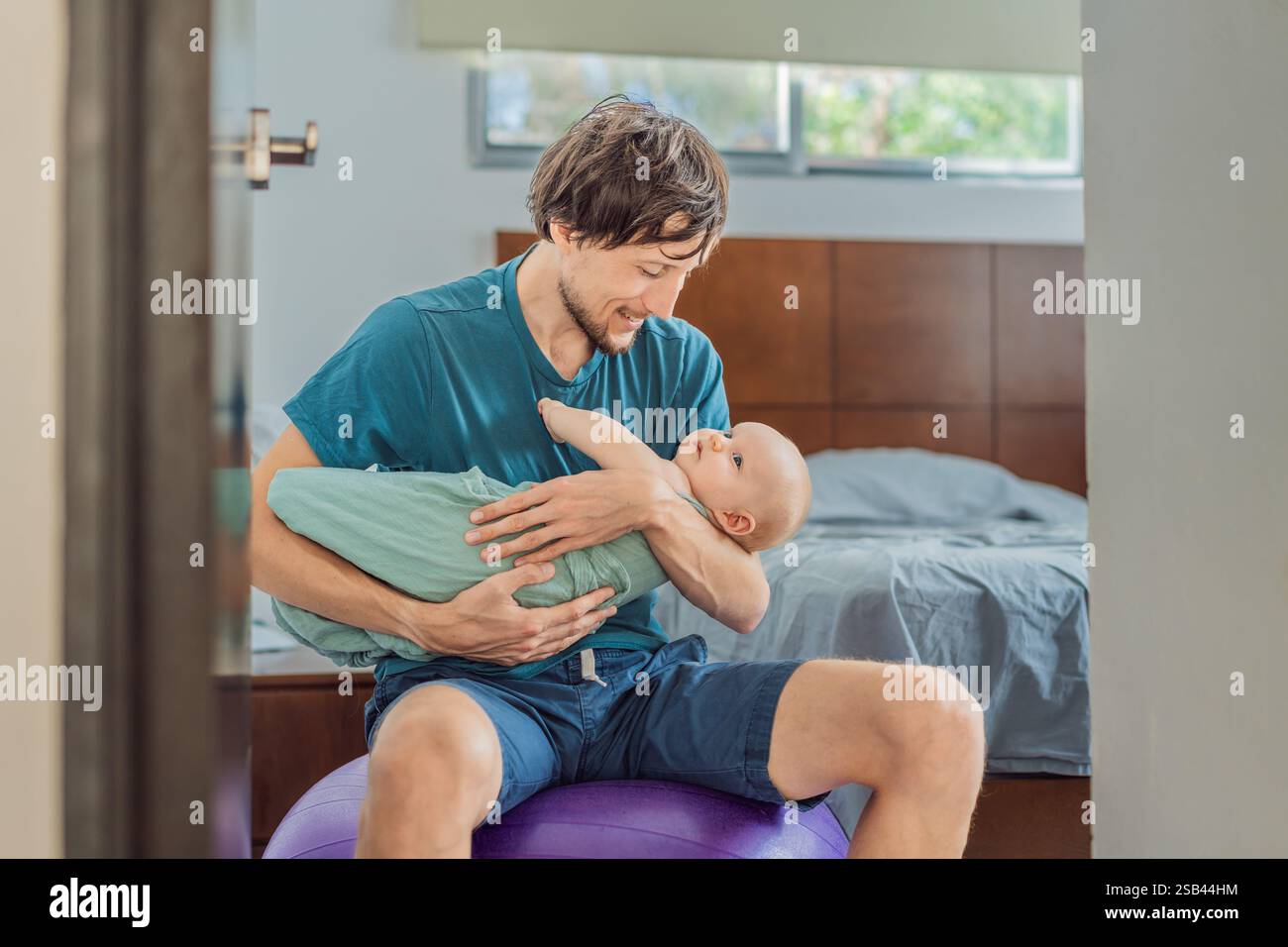A father sits on a fitness ball in a cozy bedroom, gently rocking his ...