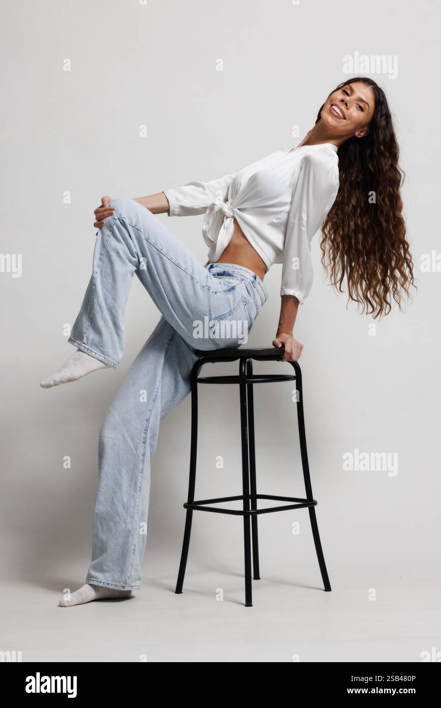 A cheerful woman posing while seated on a stool, wearing a casual ...